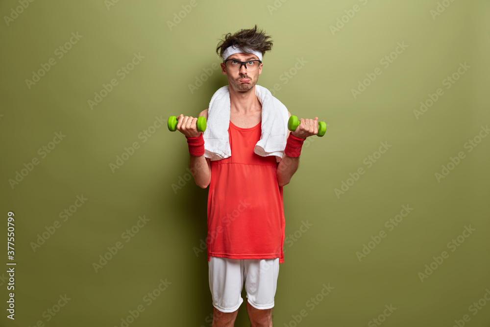 Unhappy man leads active sporty lifestyle, raises dumbbells and looks ...