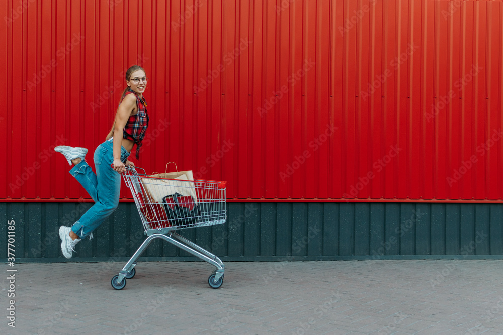 Happy girl with shopping cart on red wall shop background. Young woman ...