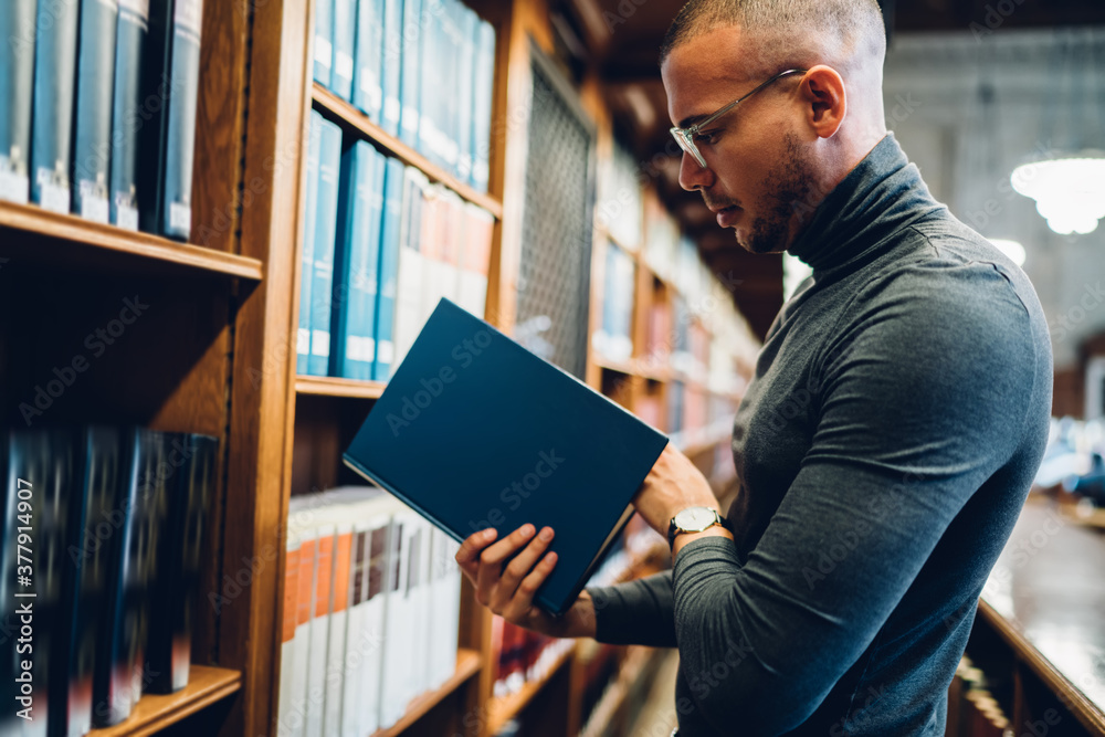 Caucasian male student choosing book for reading standing near shelves ...
