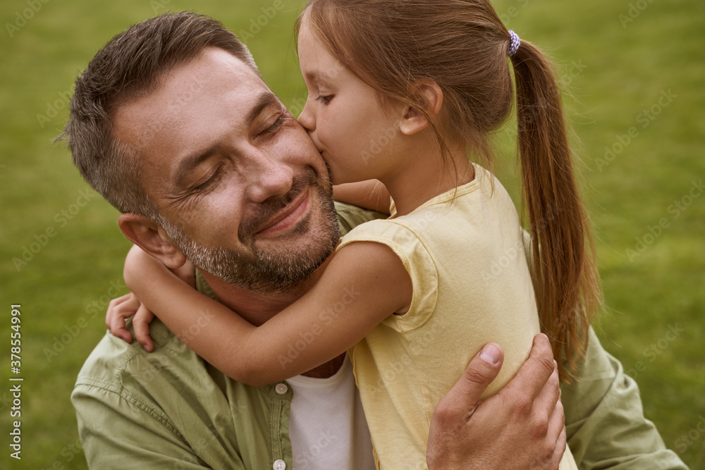 Fathers day. Cute little girl hugging and kissing her happy dad while sitting on a green grass ...