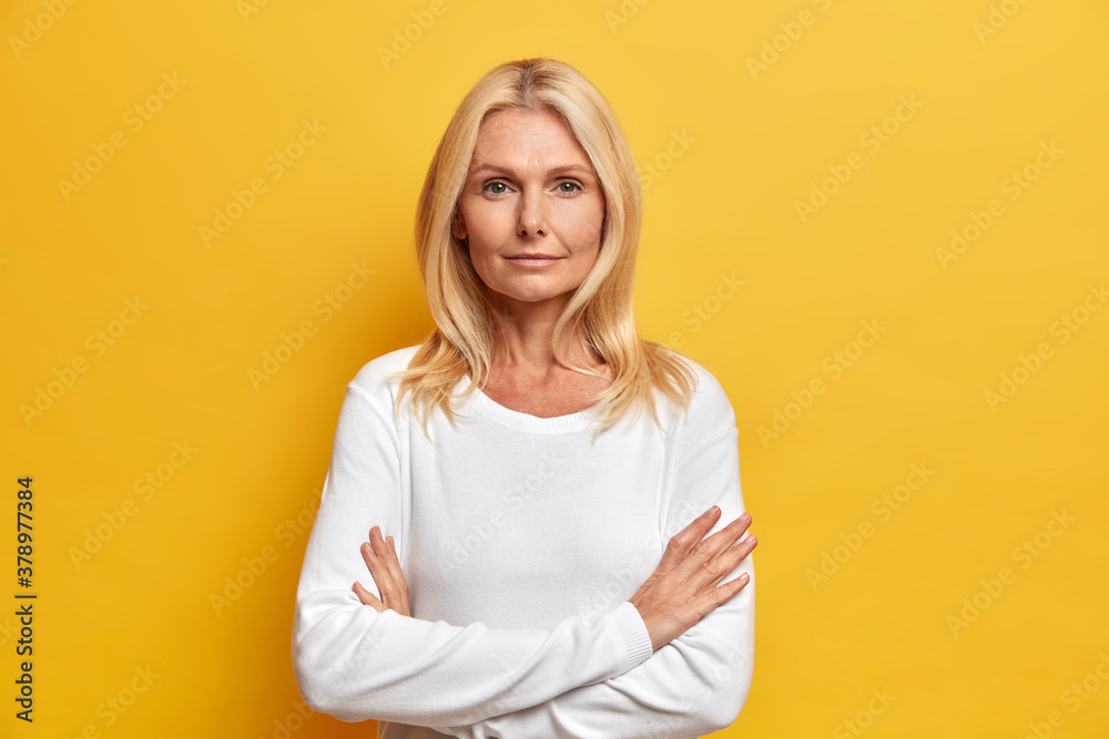 Charming serious middle aged blonde woman stands crossed hands indoor ...