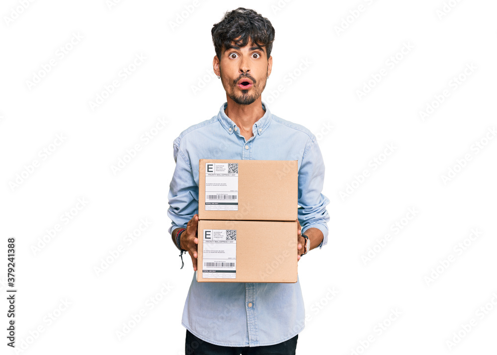 Young hispanic man holding delivery package scared and amazed with open ...