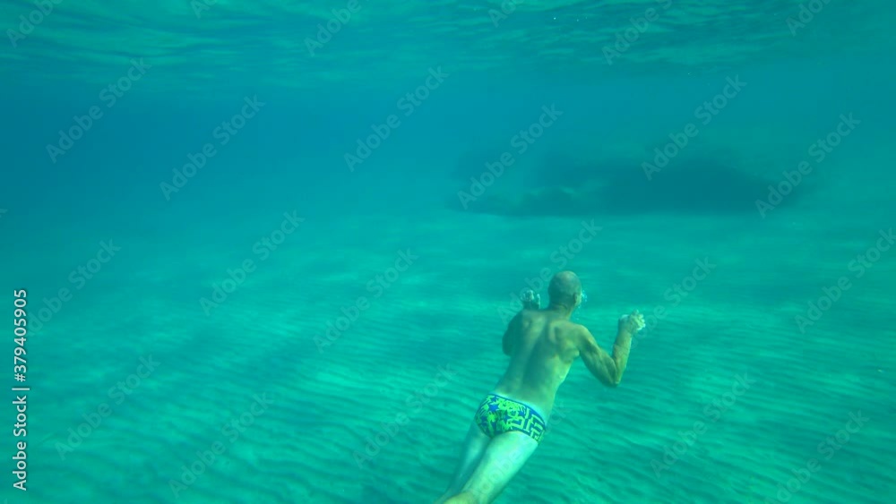 A young bald man dives into the sea under water, plunges into the sandy ...