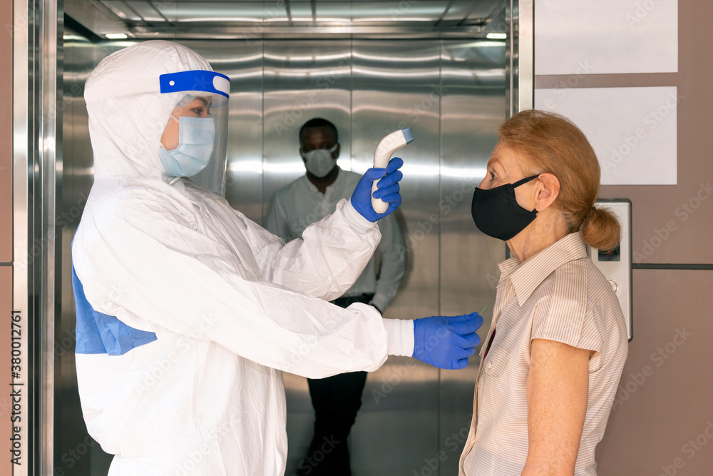 A medical worker in PPE suits using thermometer to check the employee ...
