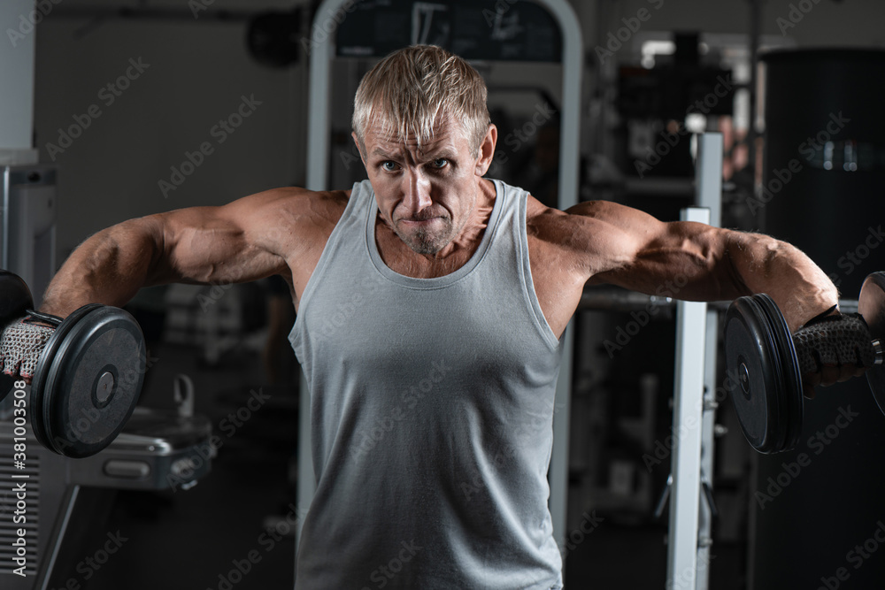 Muscular adult brutal man with dumbbells pumps biceps in the gym ...