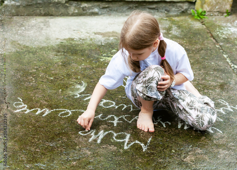 Small young girl writing simple words on concrete with white chalk ...
