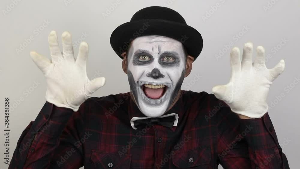 A horrible man in clown makeup grimaces and makes frightening gestures ...