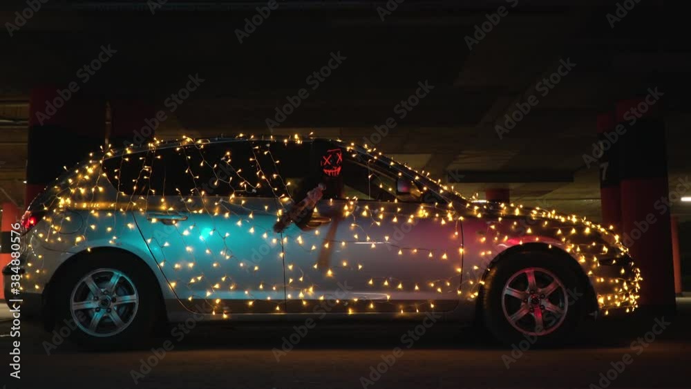 A man in a neon mask looks out of a car window in a parking lot at ...