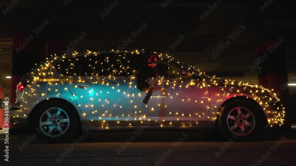 A man in a neon mask looks out of a car window in a parking lot at ...