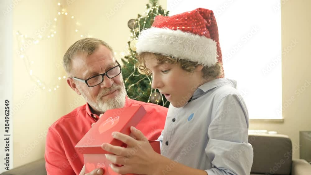 Grandpa wearing a Santa hat makes a Christmas present to his teenage ...