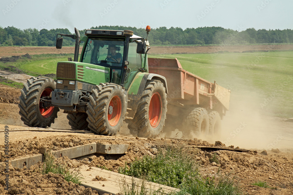A tractor with empty tipper trailer drives up a dike on a dusty track ...