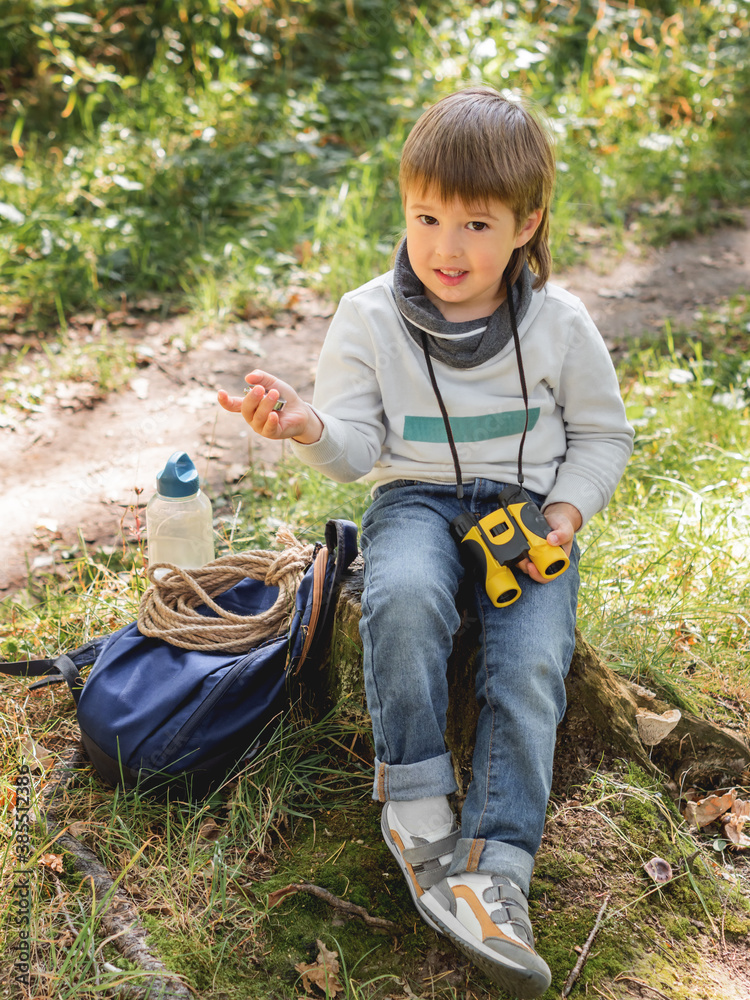 Little explorer on hike in forest. Boy with binoculars and compass sits ...