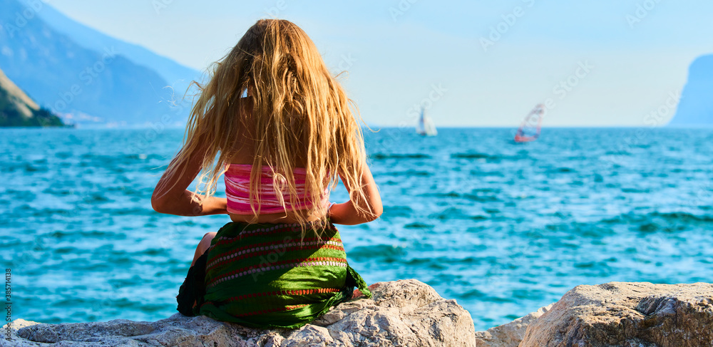 Child girl sitting on a stone on the beach. In the background lake ...