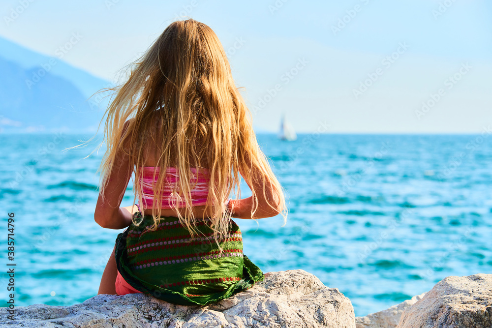 Child girl sitting on a stone on the beach. In the background lake ...