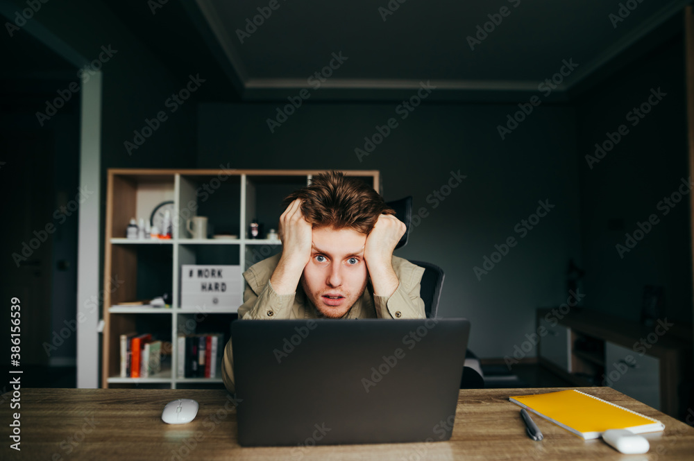 Sad young freelancer man sitting at work at home in bedroom with laptop ...