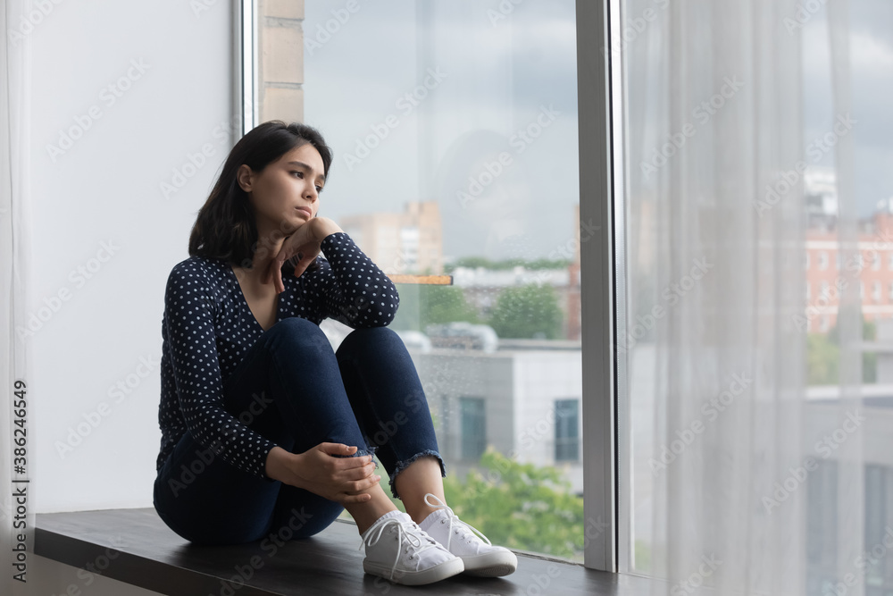 Sad young asian woman sit on windowsill at home look in window distance ...