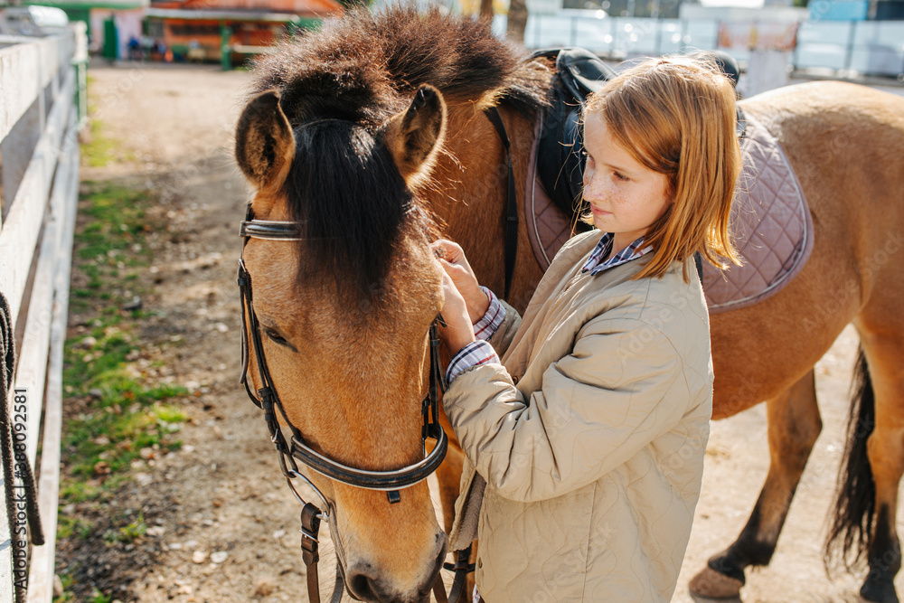 Lovely ginger girl preparing her horse for the ride, putting on bridle ...