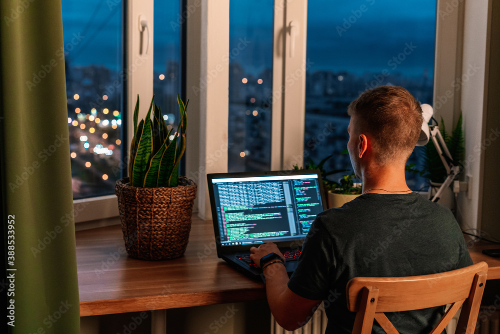 A young male programmer in front of a window with a lights of night ...