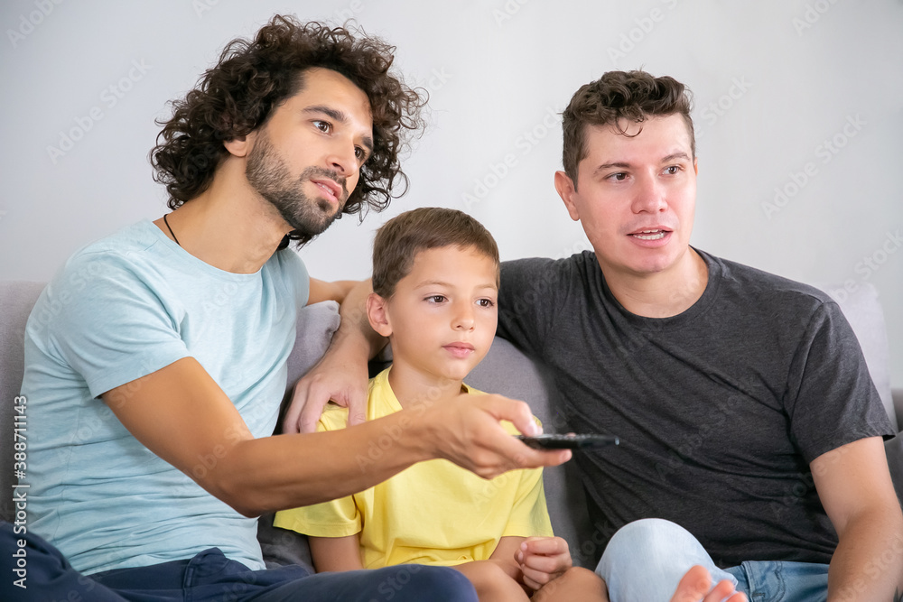 Two excited dads and son watching TV show at home, sitting on couch in ...