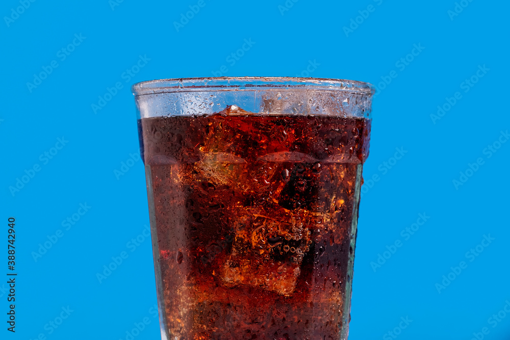 Ice cold glass of soda, cola, fizzy drink, carbonated beverage sits against a blue wall with ice ...