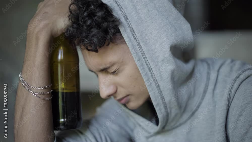 Close-up of drunk depressed teenager in hood holding beer bottle and ...
