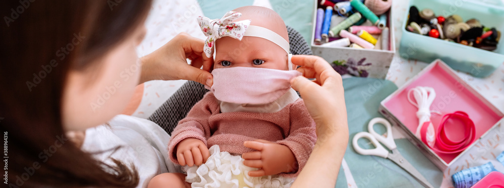 Little girl trying on her baby doll a cloth mask that she is sewing ...