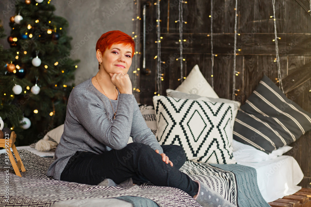 Red-hair lady weared in knitted sweater in Christmas decorated room with garland lights sitting ...