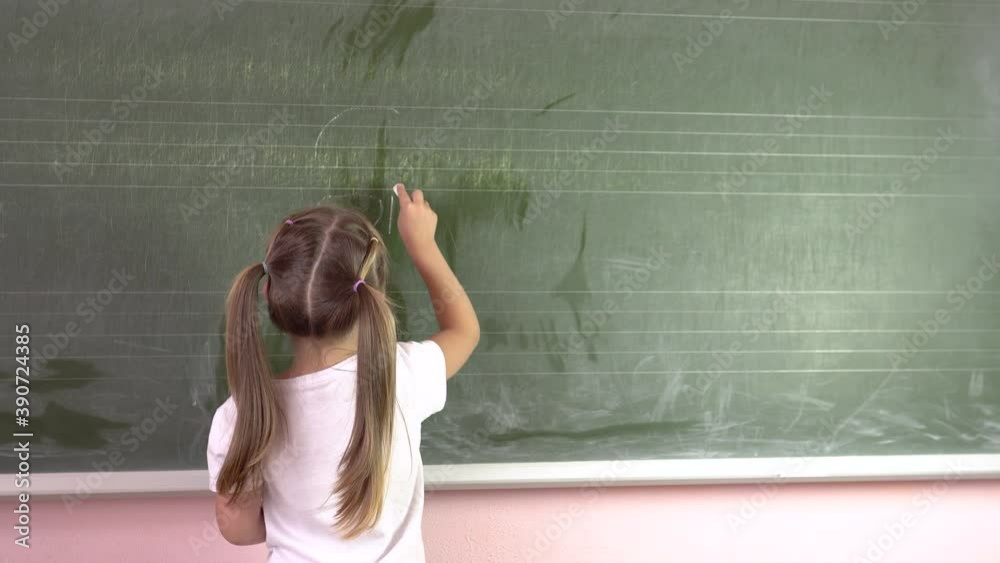 Little girl student writes letters in chalk on a school board ...