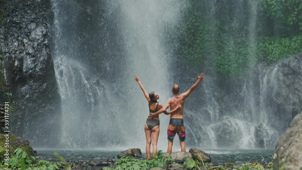 Rear view of young traveling couple standing in front of waterfall with ...