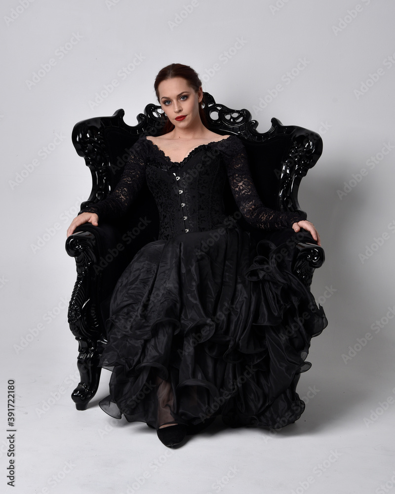 full length portrait of woman wearing black gothic dress, sitting on a ...