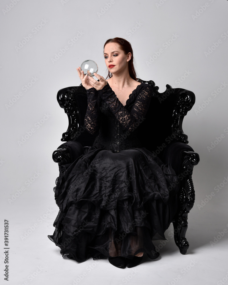 full length portrait of woman wearing black gothic dress, sitting on a ...