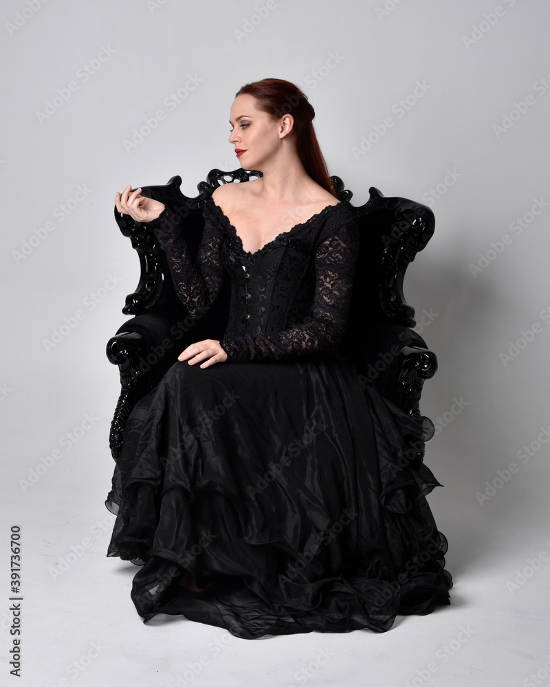 full length portrait of woman wearing black gothic dress, sitting on a ...