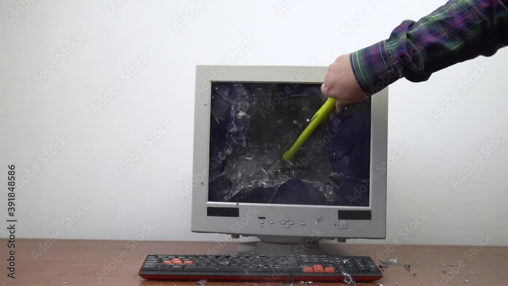 An old monitor sitting on a table is smashed with a hammer. The ...