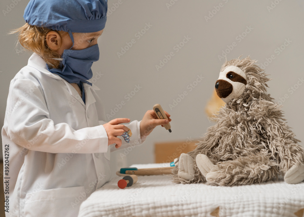 young child in medical PPE examines a plush toy sloth and takes it's ...