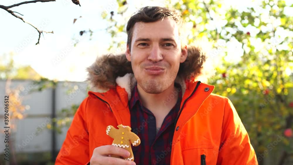 Young man in orange jacket bites gingerbread man, standing in garden ...