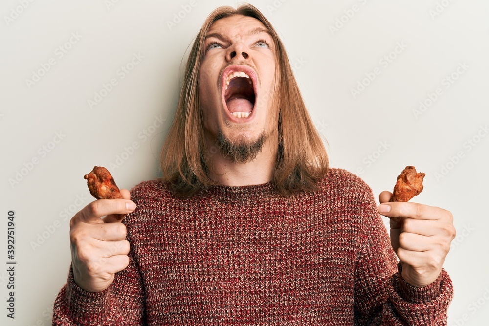 Handsome caucasian man with long hair eating chicken wings angry and ...
