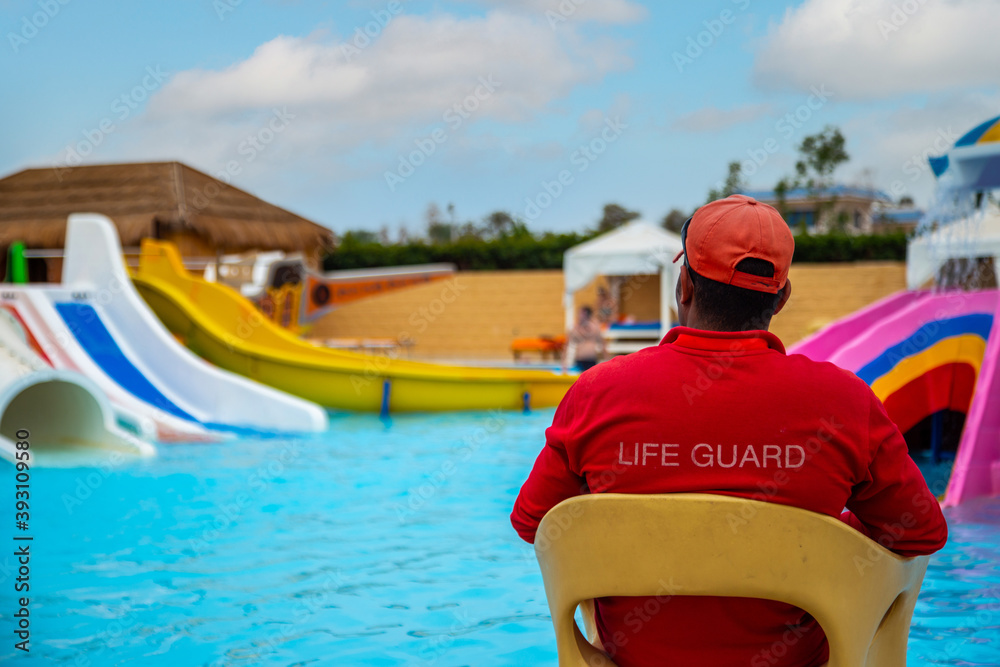 Rear view of lifeguard sitting on chair with rescue buoy at poolside ...