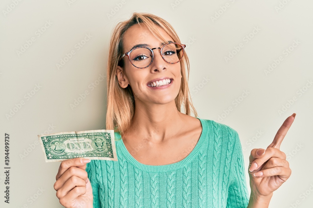 Beautiful blonde woman holding 1 dollar banknote smiling happy pointing ...