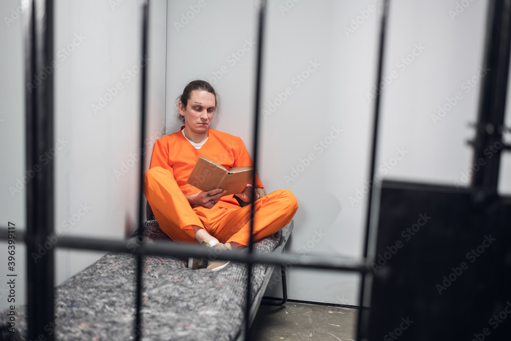 Young criminal in a solitary cell reads a book sitting on a bunk in an ...