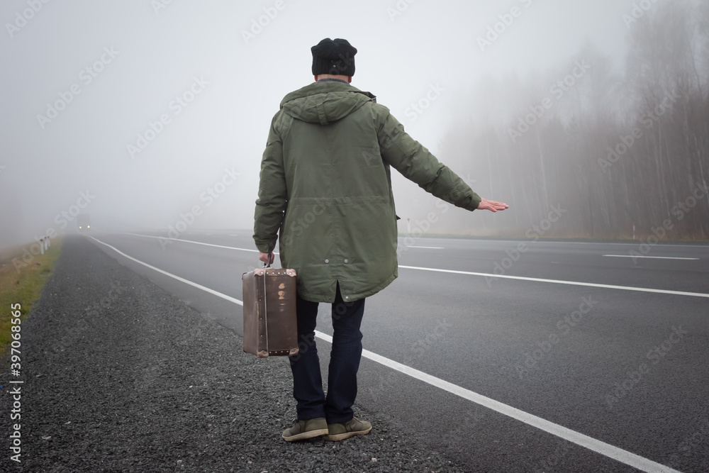 A young man with a suitcase in his hands stands on the side of the road ...