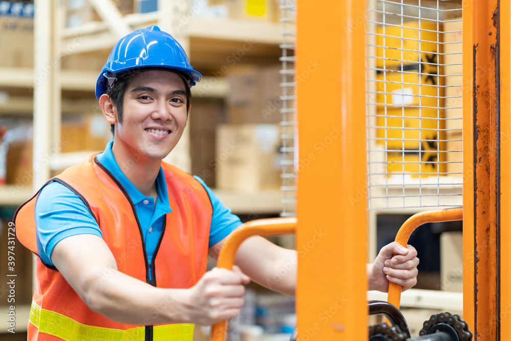 Smiling Asian male warehouse worker in safety vest and helmet driving and operating on forklift ...