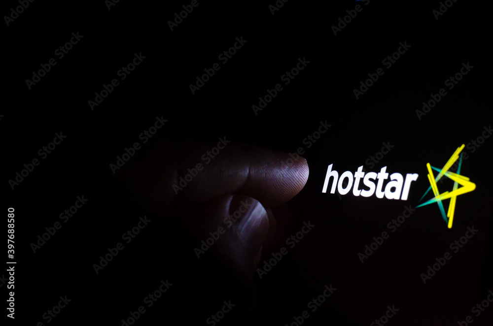 Hotstar streaming service logo on the screen in a dark room and a hand ...