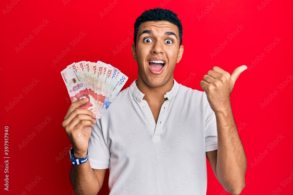 Young arab man holding colombian pesos pointing thumb up to the side ...
