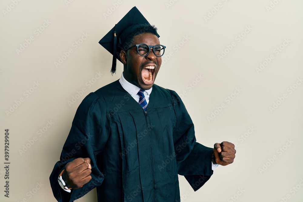 Handsome black man wearing graduation cap and ceremony robe angry and ...