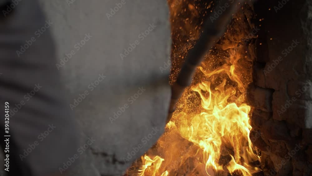 A dark skinned worker poking and adjusting a fire with a wooden stick ...