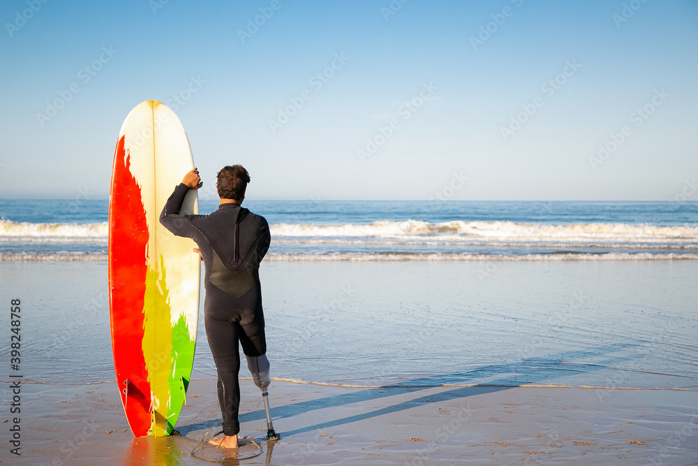 Back view of brunette surfer standing with surfboard on beach. Disabled ...