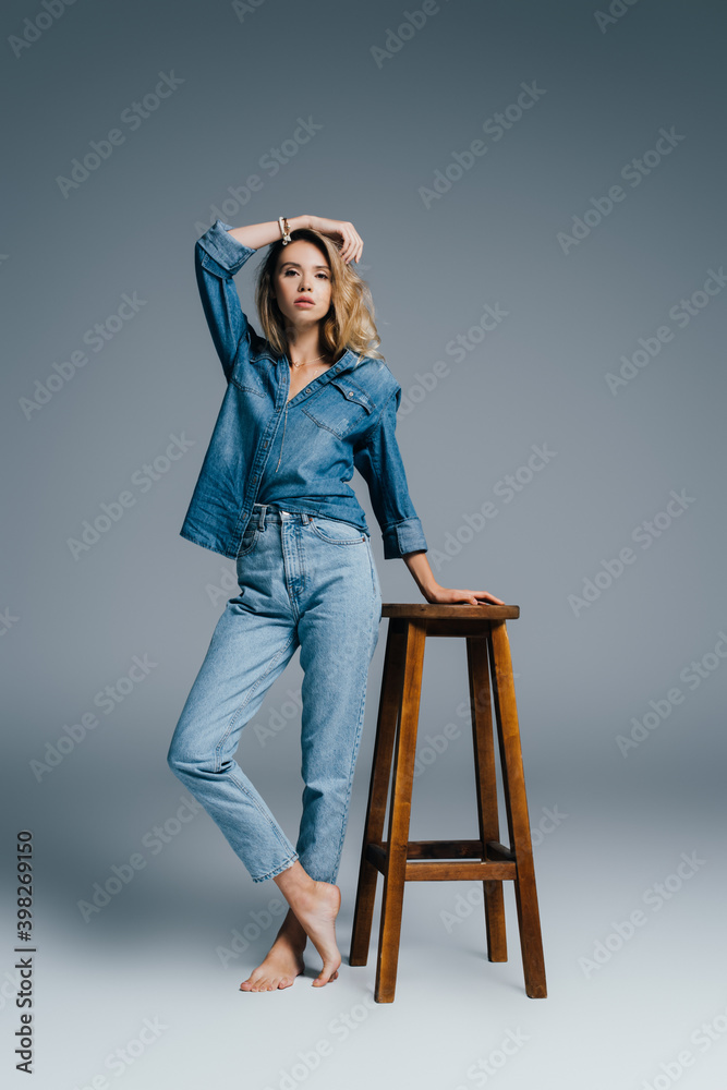stylish barefoot woman in denim shirt and jeans leaning on high stool ...