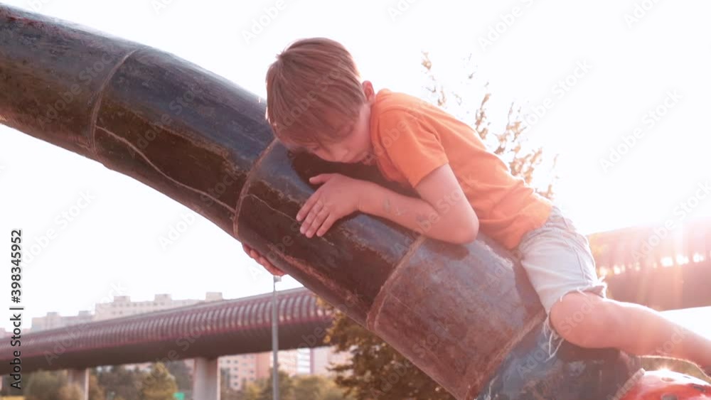 A boy climbs a pipe installed on a playground in the rays of the summer ...