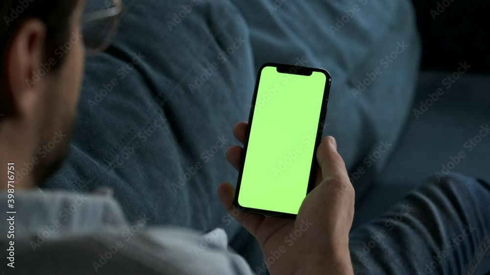 Man lying on couch using smartphone with chroma key green screen at night, scrdoing various ...