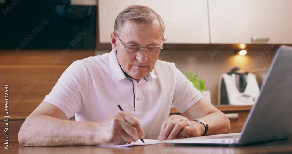 An older man writes his notes onto paper from his laptop. Grandfather ...